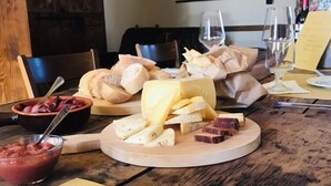 Uma mesa rústica com diversos queijos, pão, uma tigela de frutas e uma taça de vinho.