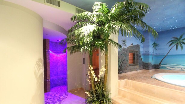 Uma área de piscina interior luxuosa com tema tropical, incluindo uma palmeira e um mural com uma cena de praia.