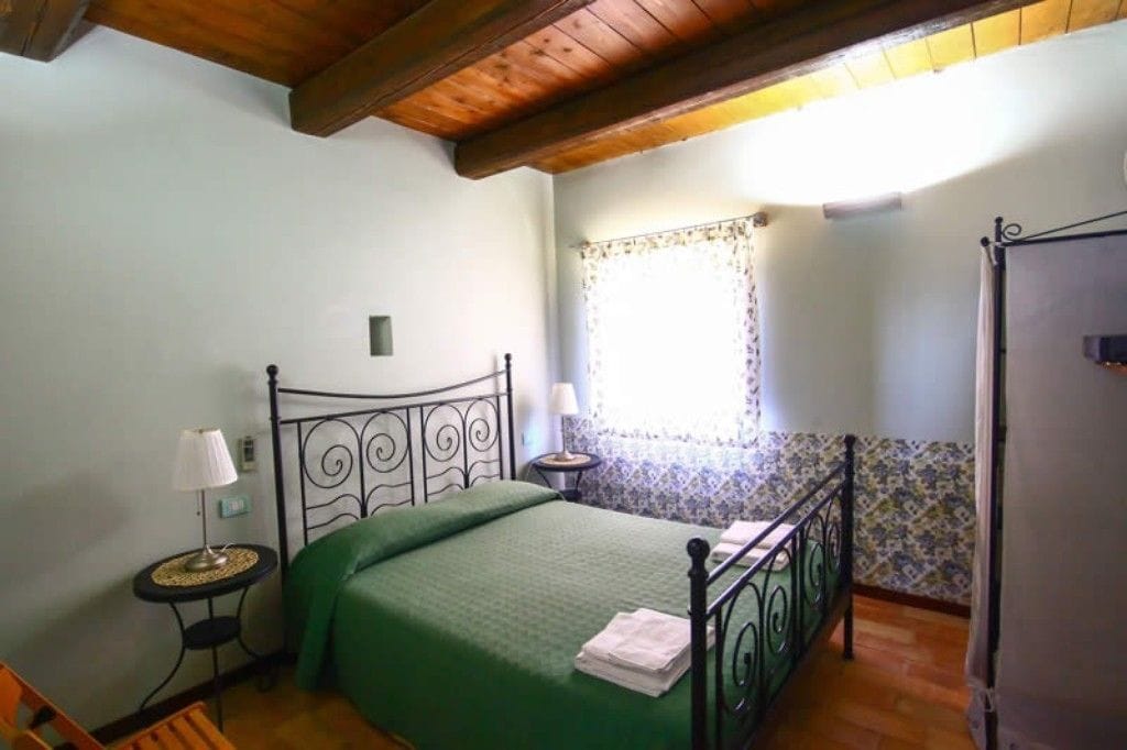 Quarto com cama, mesa de cabeceira, abajur, janela com cortinas e teto de madeira.