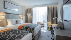 Quarto de hotel com duas camas, uma escrivaninha, uma cadeira, um abajur e uma janela grande com cortinas.