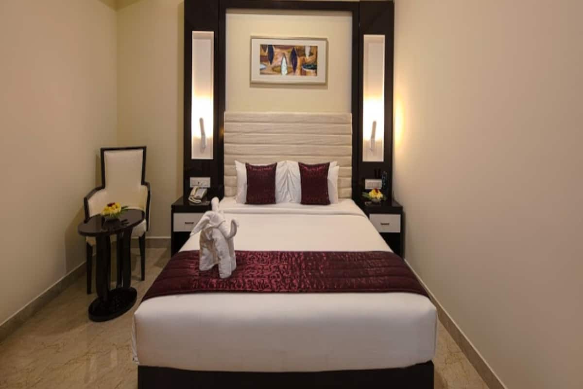 Quarto de hotel com uma cama grande, mesa de cabeceira com abajur, uma cadeira e uma mesinha com um vaso de flores.