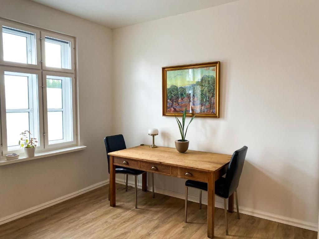 Uma sala de jantar com mesa de madeira, quatro cadeiras, planta em vaso, luminária e pintura na parede.