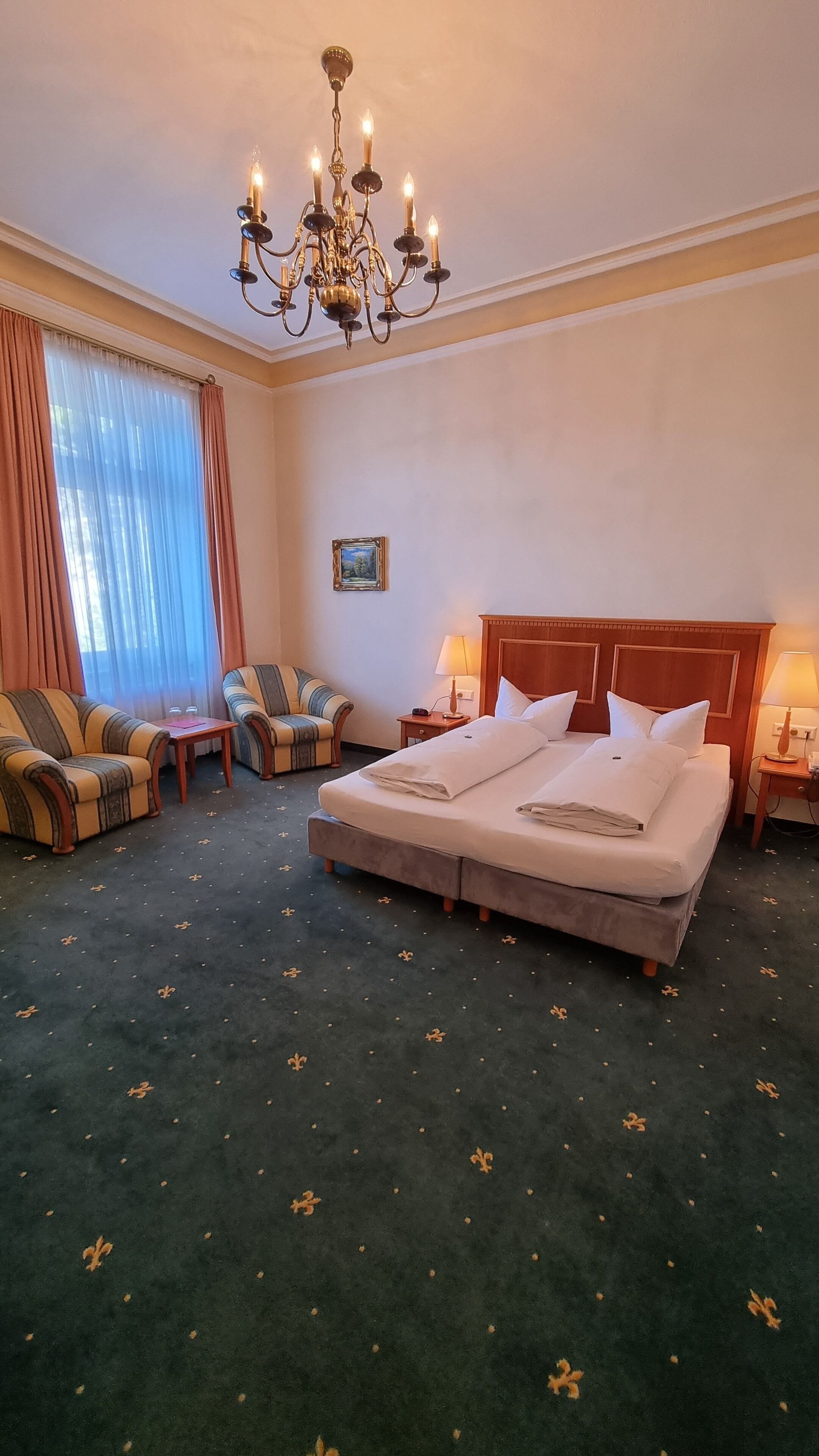 Quarto de hotel com uma cama grande, duas poltronas, um candelabro e vista para o exterior.
