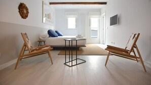 Quarto com cama, duas cadeiras de madeira, uma mesinha e uma TV na parede.