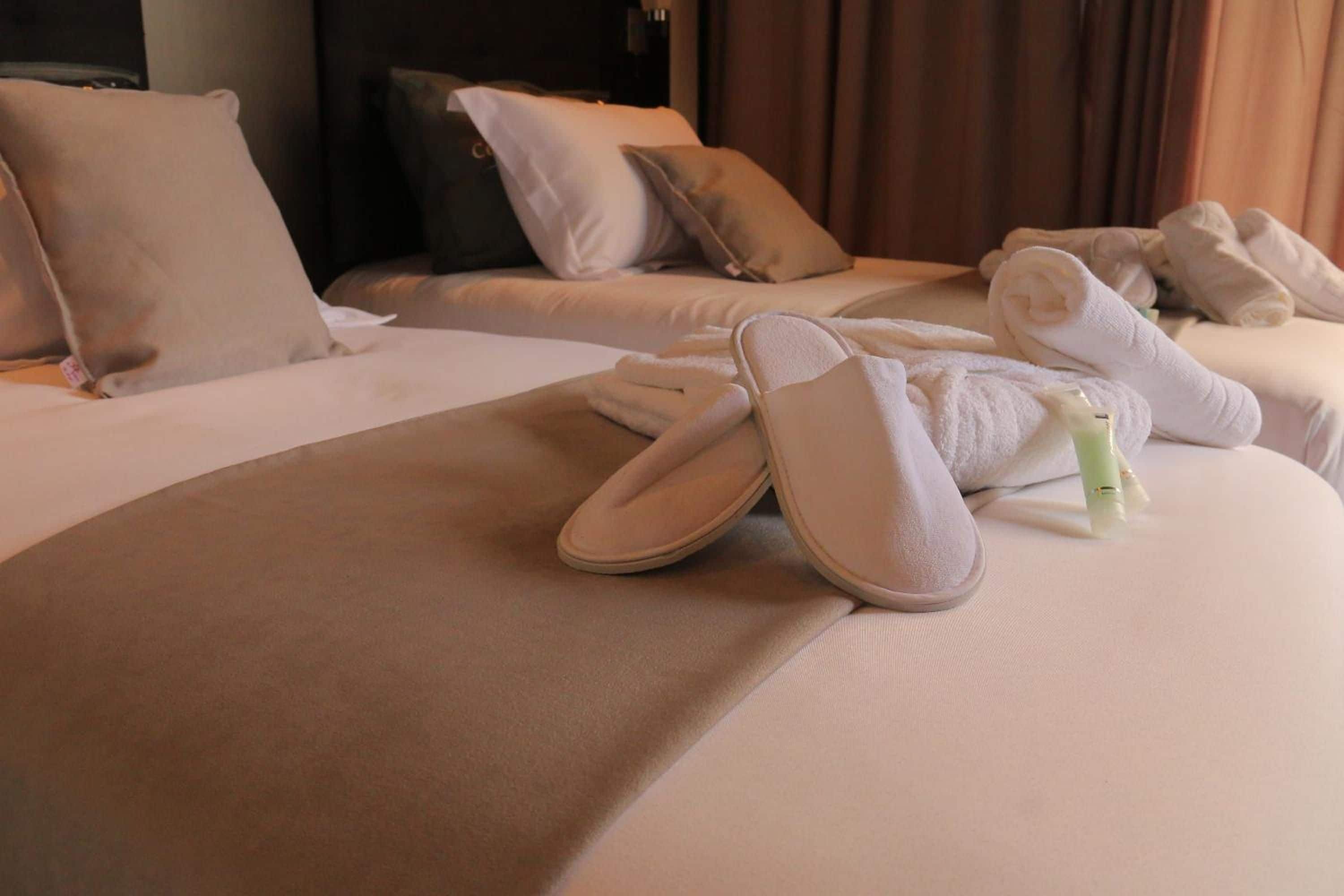 Quarto de hotel com cama bem arrumada, chinelos e um frasco de loção sobre a cama.