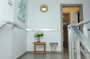 Um corredor com uma mesa consola de madeira, um vaso de flores brancas, um espelho redondo e um banquinho estofado verde.