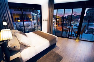 Quarto de hotel moderno com uma cama grande, uma mesa de cabeceira e vista para o horizonte urbano através de janelas do chão ao teto.