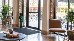 Um lobby espaçoso com interior moderno, apresentando uma área de estar com sofá e cadeiras, uma mesa de centro com livros, e grandes janelas com vista para a rua.