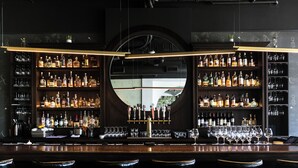 Um balcão de bar com prateleiras cheias de garrafas de bebidas e copos, e luminárias pendentes.