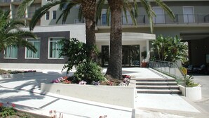 Entrada de hotel com uma palmeira, escadaria e um edifício com varandas.