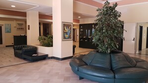 Um saguão com balcão de atendimento, área de recepção, espaço de estar com sofá e poltrona, e um vaso de plantas.
