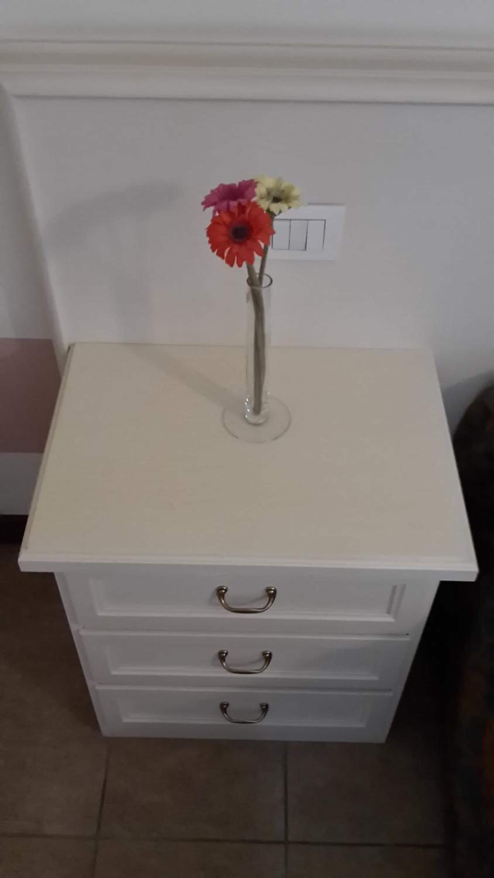 Uma mesa de cabeceira branca com um vaso contendo três flores e três gavetas com puxadores de metal.