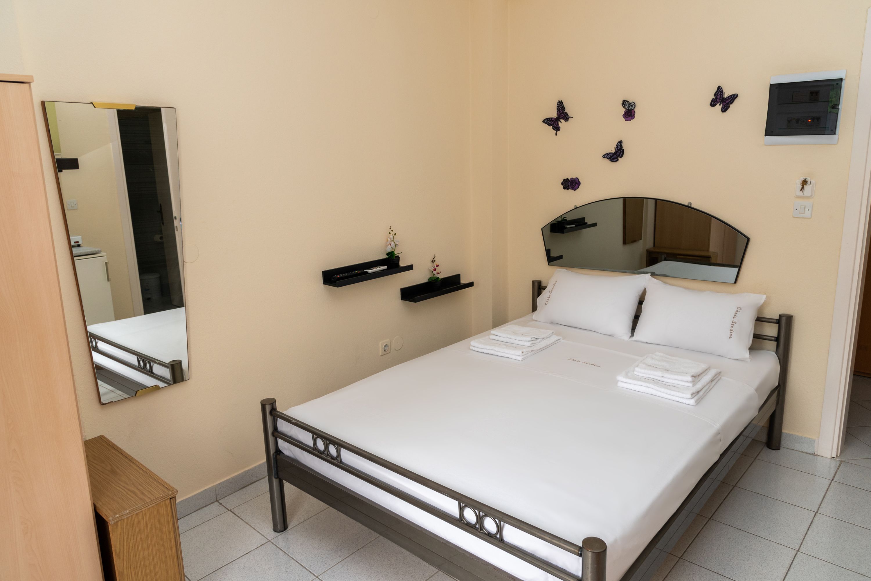 Um quarto bem arrumado com cama, mesa de cabeceira, espelho e prateleiras na parede com objetos decorativos.
