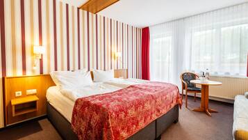 Quarto de hotel com uma cama grande, uma mesa de cabeceira, uma cadeira, uma mesinha redonda e uma janela com cortinas.