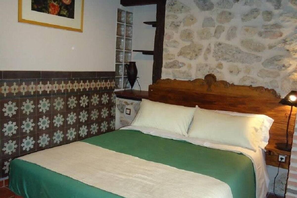 Quarto com parede de pedra, cabeceira de madeira, cama com roupa de cama verde e branca, quadro na parede e pequena mesa com abajur.