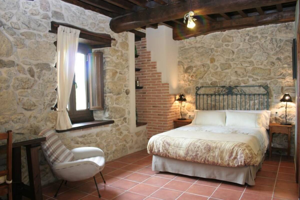 Quarto com parede de pedra, cama com colcha estampada, uma cadeira, janela com cortinas e escada.