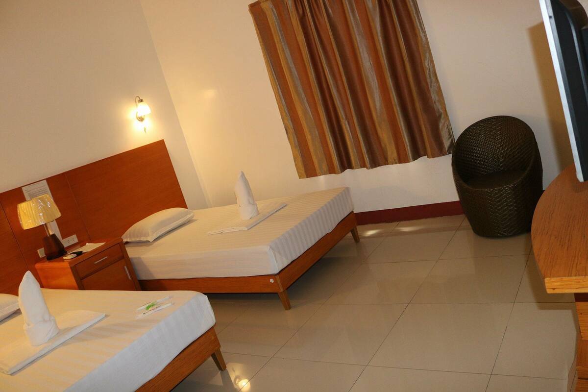 Quarto de hotel com duas camas de solteiro, cabeceira de madeira, mesa de cabeceira com luminária, lixeira e cortina.