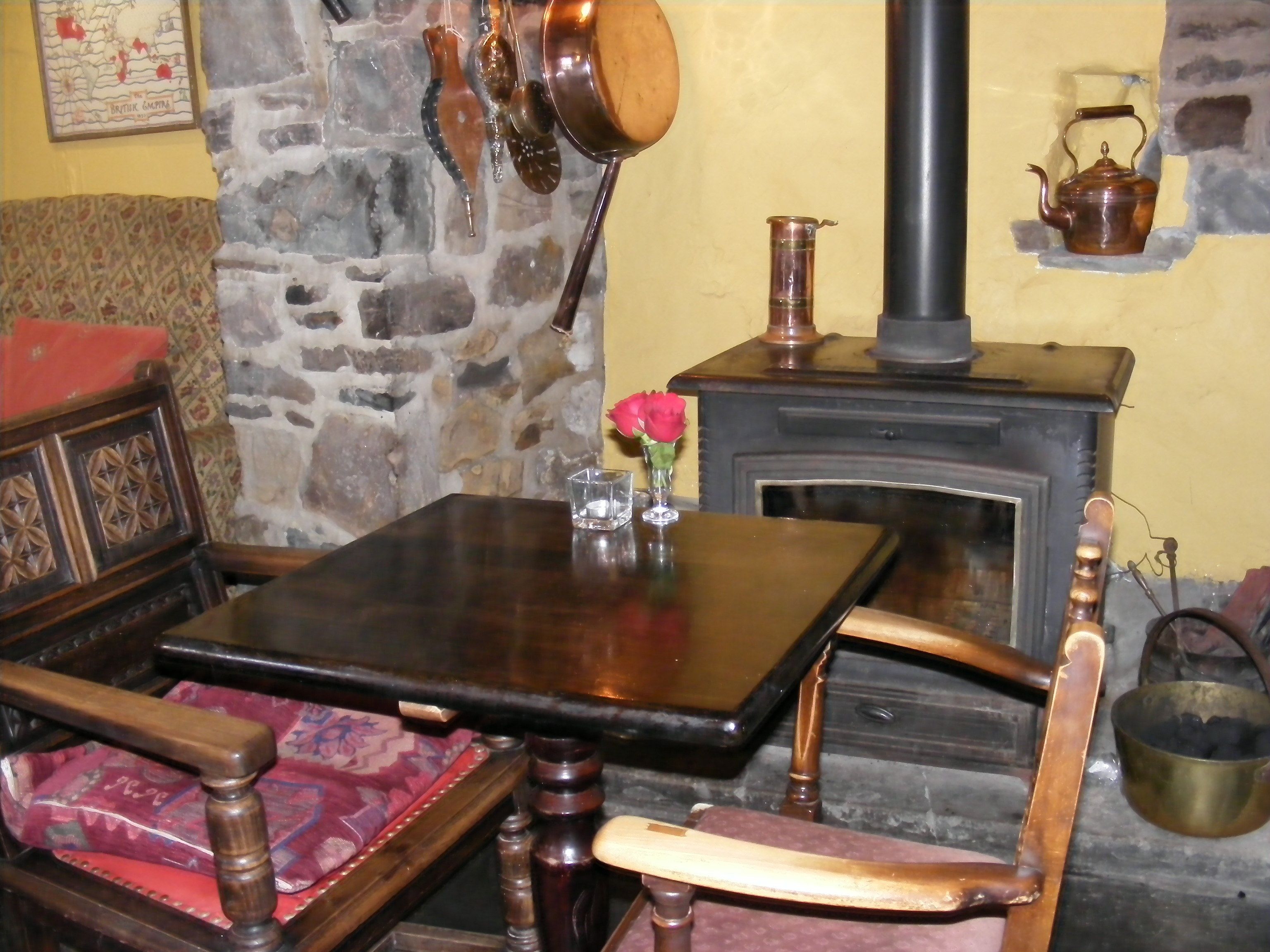Quarto tradicional com parede de pedra, uma mesa de madeira, uma cadeira, um pequeno vaso com uma flor e uma lareira com um bule.