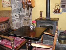 Quarto tradicional com parede de pedra, uma mesa de madeira, uma cadeira, um pequeno vaso com uma flor e uma lareira com um bule.