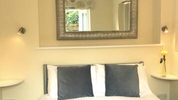 Cama bem arrumada com duas almofadas, mesa de cabeceira com um vaso de flores e um espelho sobre a cama.