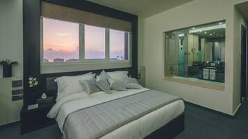 Quarto de hotel moderno com uma cama grande, uma janela com vista para a cidade, um espelho e uma mesinha de cabeceira.