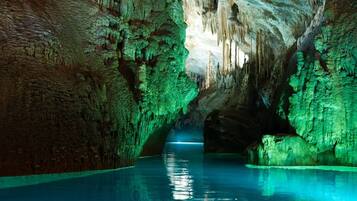 Caverna com estalactites e estalagmites, iluminada por luzes verdes, e um lago de água no fundo.