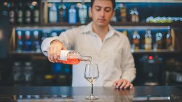 Um barman servindo uma bebida num copo num bar, com prateleiras cheias de garrafas ao fundo.