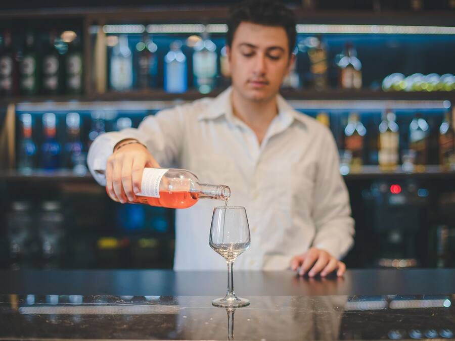 Um barman servindo uma bebida num copo num bar, com prateleiras cheias de garrafas ao fundo.