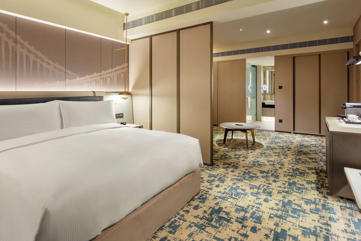 Quarto de hotel com uma cama grande, uma pequena mesa redonda, uma secretária com cadeira e um banheiro visível através de uma porta de vidro.