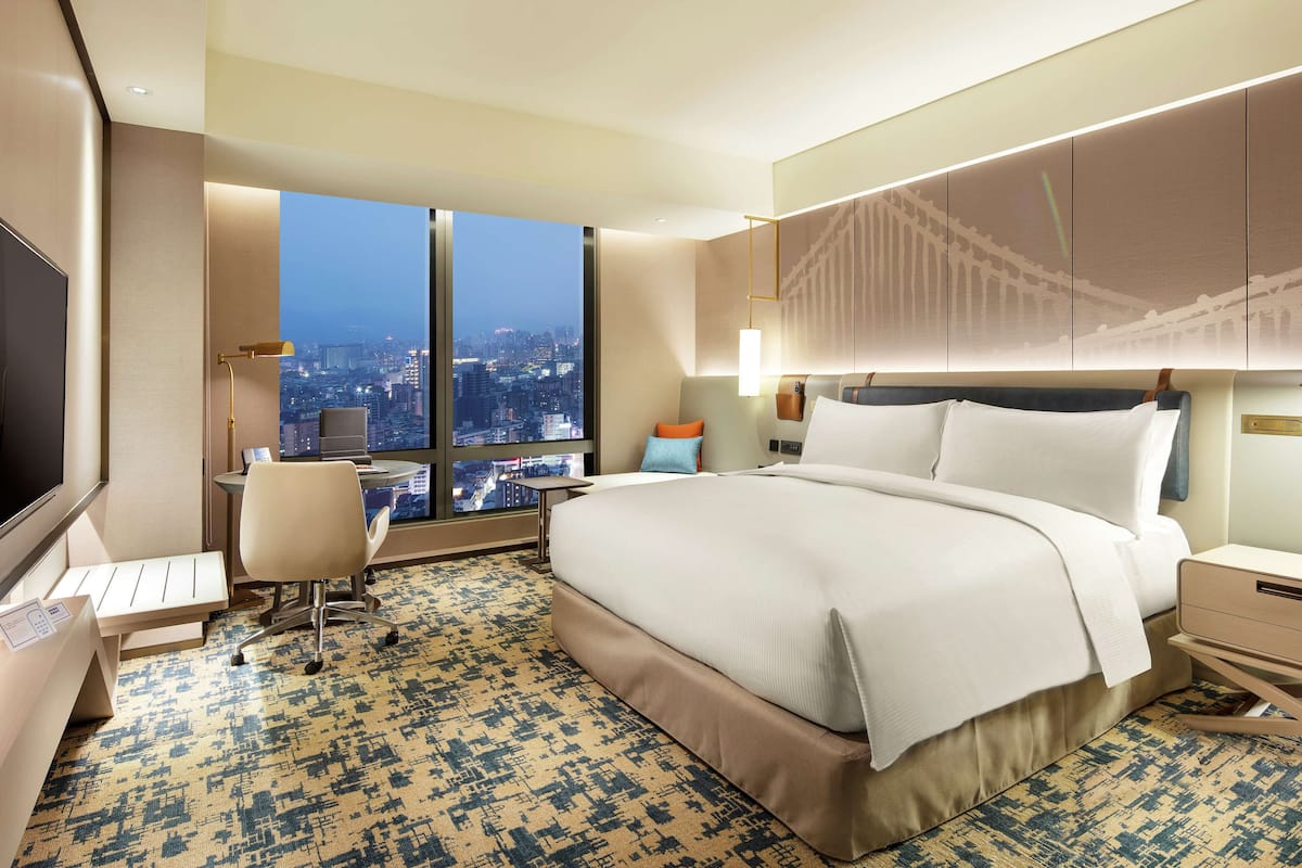 Quarto de hotel com uma cama grande, uma escrivaninha, uma cadeira e vista para a paisagem urbana através de amplas janelas.