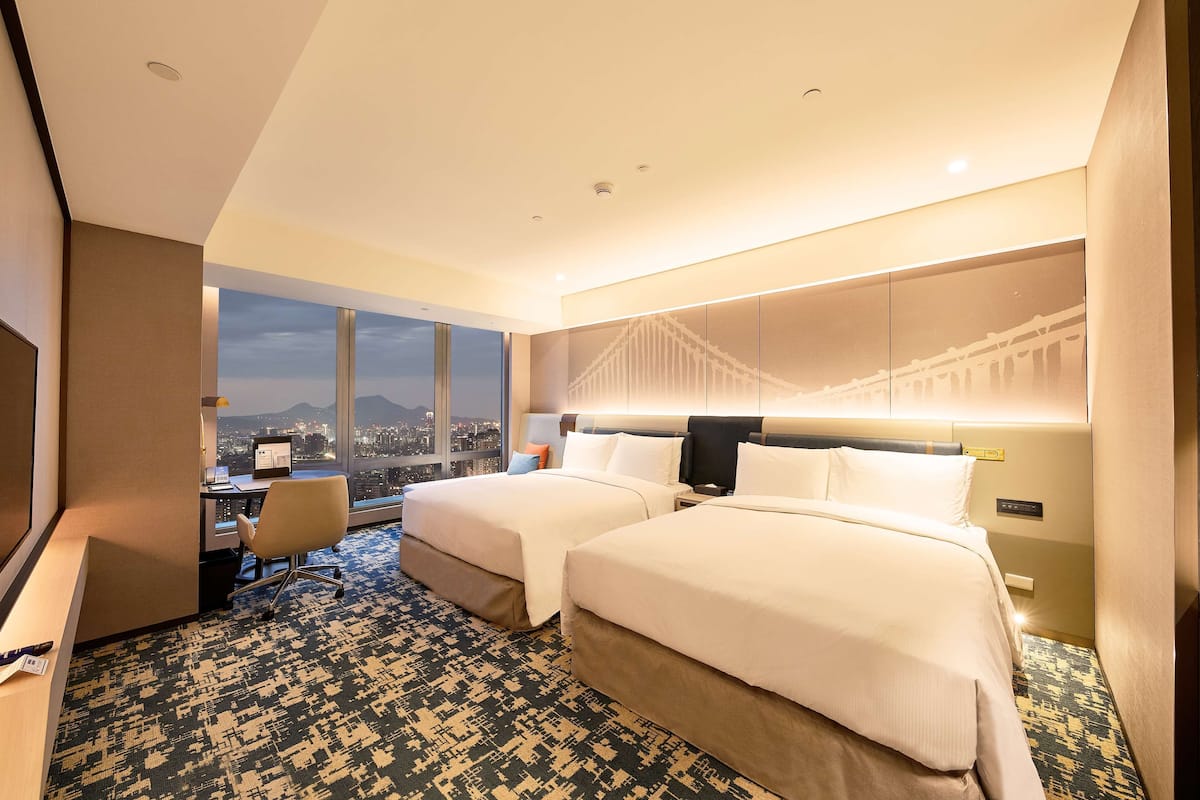 Quarto de hotel com duas camas, uma escrivaninha e uma janela grande com vista para a cidade.