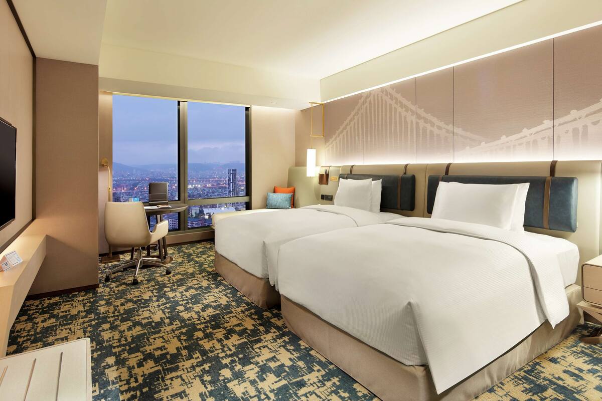 Quarto de hotel com duas camas, uma escrivaninha, uma cadeira e uma janela grande com vista para a cidade.