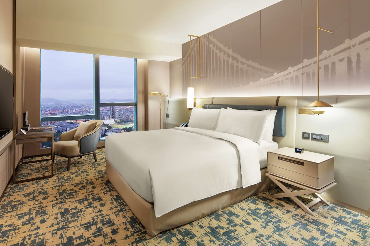 Quarto de hotel com uma cama grande, uma escrivaninha, uma cadeira e vista para a cidade através de uma grande janela.