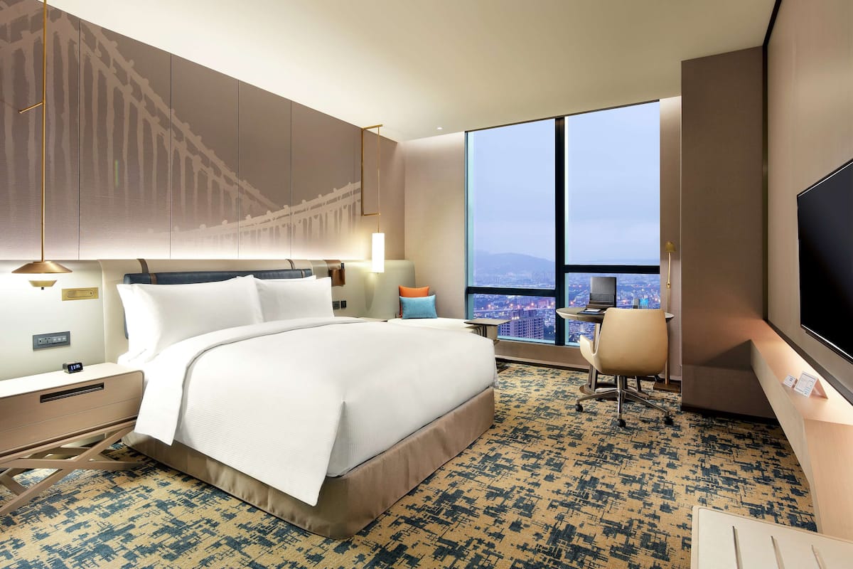 Quarto de hotel moderno com uma cama grande, uma escrivaninha com um laptop, uma TV e vista para a cidade através de amplas janelas.