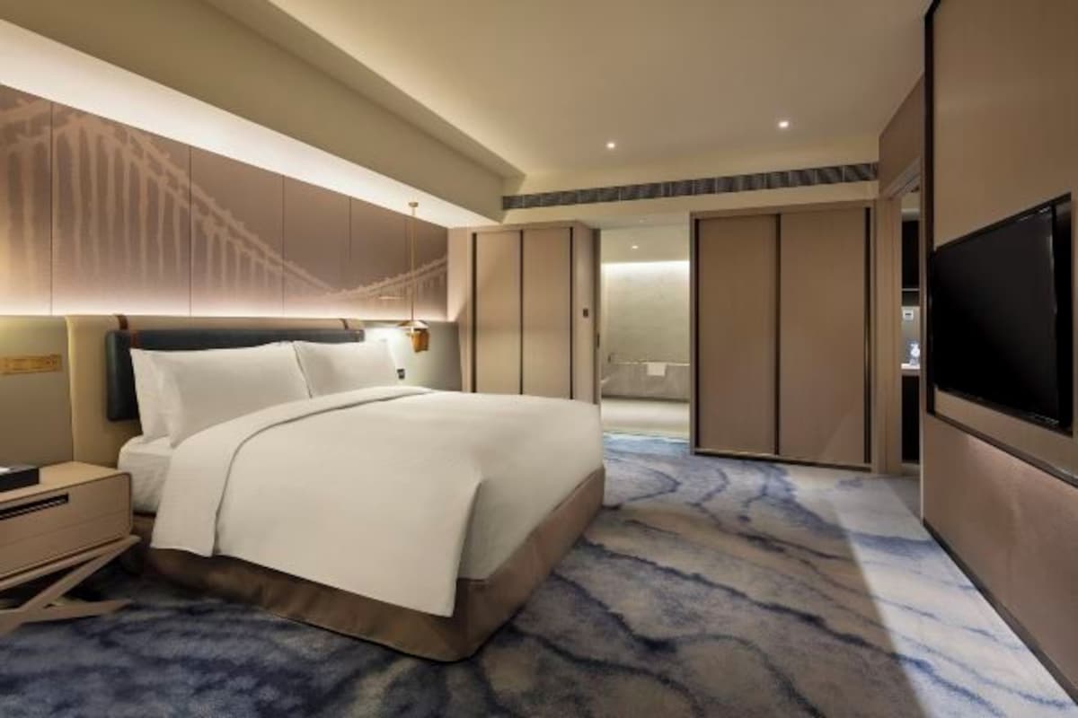 Quarto de hotel moderno com uma cama grande, televisão de tela plana, guarda-roupa e banheiro visível através de uma porta aberta.