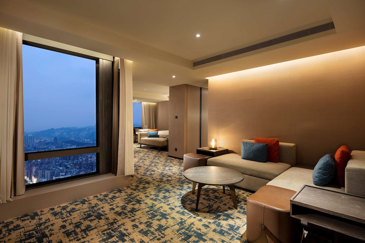 Quarto de hotel moderno com uma grande janela que oferece vista da cidade, um conjunto de sofás, uma mesa de centro redonda e uma pequena mesa lateral com uma luminária.