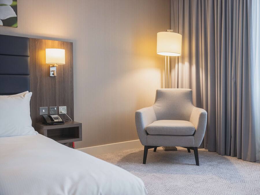 Quarto de hotel com uma cama, mesa de cabeceira com telefone, luminária e uma cadeira.
