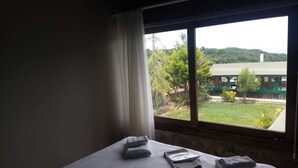 Quarto com cama, janela e vista para um edifício e vegetação lá fora.