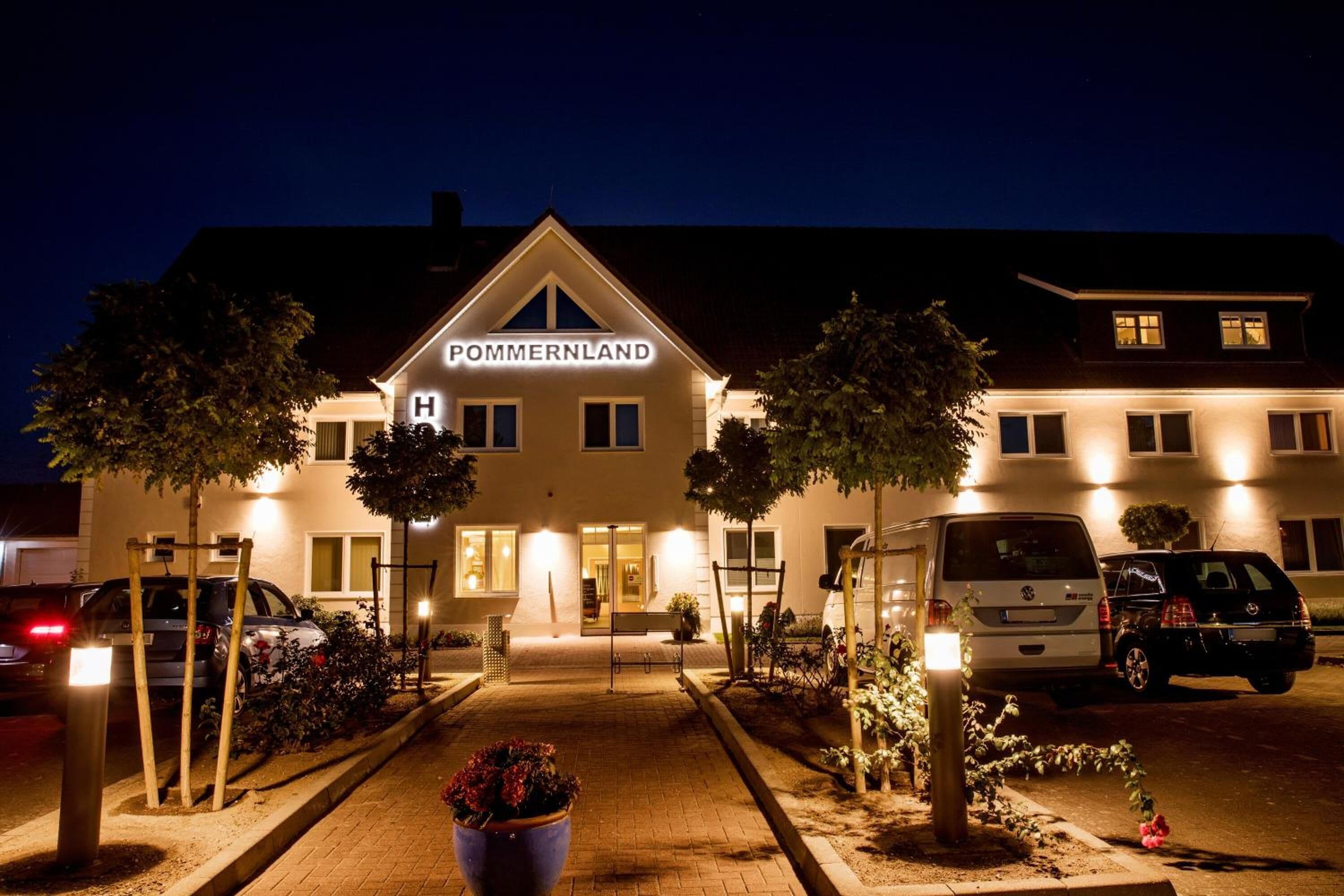 Um hotel chamado "POMMERNLAND" com entrada bem iluminada, área de estacionamento e um jardim com plantas em vasos.