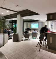 Espaço moderno em ambiente interno com piso ladrilhado, áreas de estar e uma grande tela exibindo um evento esportivo.