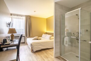 Quarto de hotel com cama, escrivaninha, cadeira e área de chuveiro envidraçada.