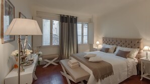 Um quarto com uma cama grande, mesas de cabeceira, um banquinho e uma escrivaninha com abajur e flores.