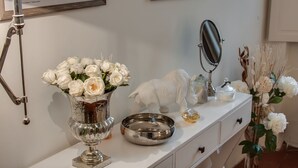 Uma mesinha de vaidade branca com espelho, um vaso de flores brancas, uma tigela de prata e um abajur.