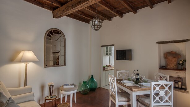 Uma área de refeições com mesa de madeira posta para quatro pessoas, cadeiras brancas e um abajur sobre uma mesinha lateral.