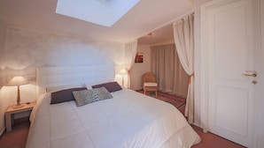 Quarto com uma cama grande, duas luminárias de cabeceira, uma cadeira, uma porta e um claraboia.