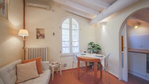 Uma sala de estar aconchegante com um sofá branco, uma mesa de jantar com cadeiras, um radiador e uma janela com vista.