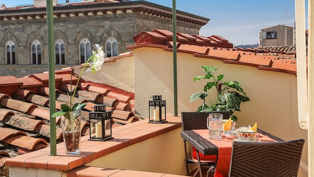 Um terraço com uma mesa posta para dois, um vaso de planta e vista para um edifício histórico com arcos.