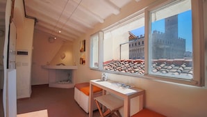 Quarto compacto com escrivaninha, um pequeno espaço para sentar e uma janela com vista para um edifício histórico.