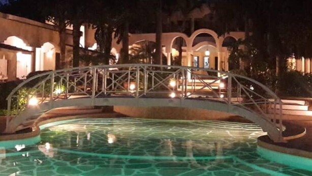 Vista noturna de uma área de piscina com uma ponte, palmeiras e um prédio com janelas em arco.