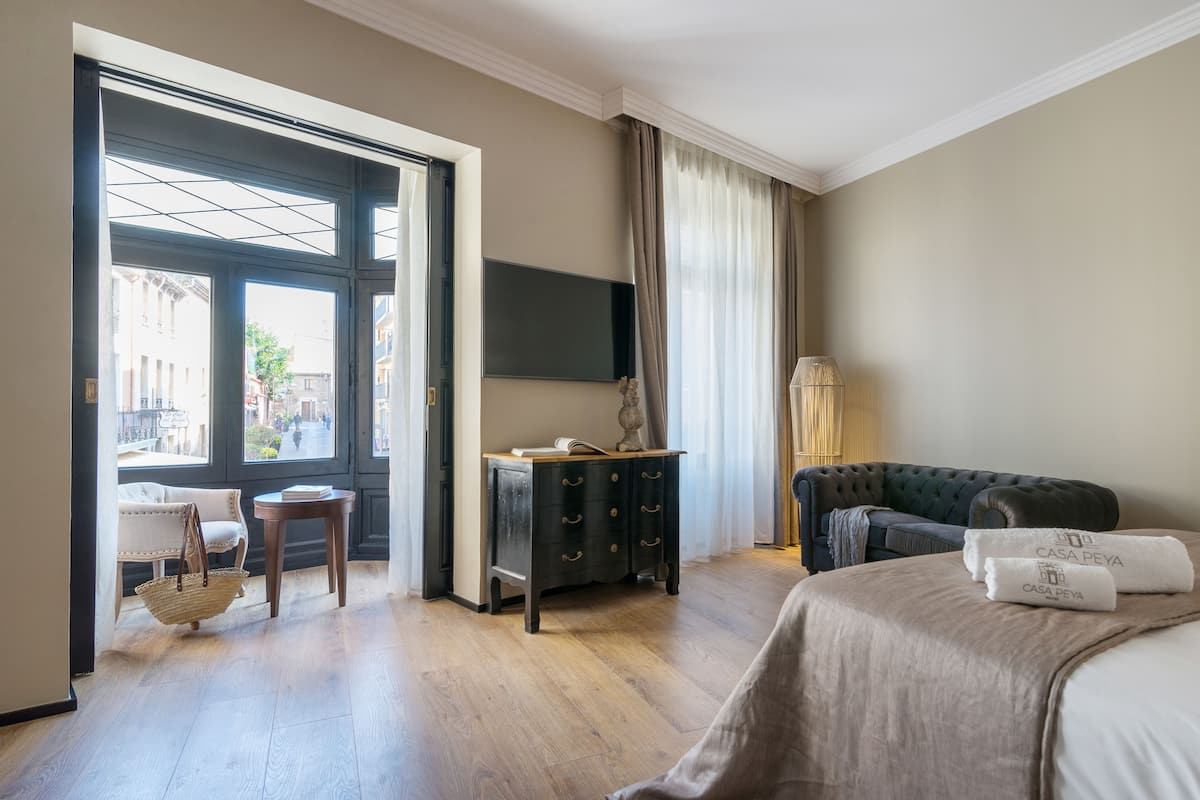 Quarto de hotel com cama, escrivaninha com luminária, cadeira, janela com cortinas e vista para a rua.